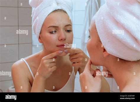 Young woman looking and squeeze acne on a face in front of the mirror. Ugly problem skin girl ...